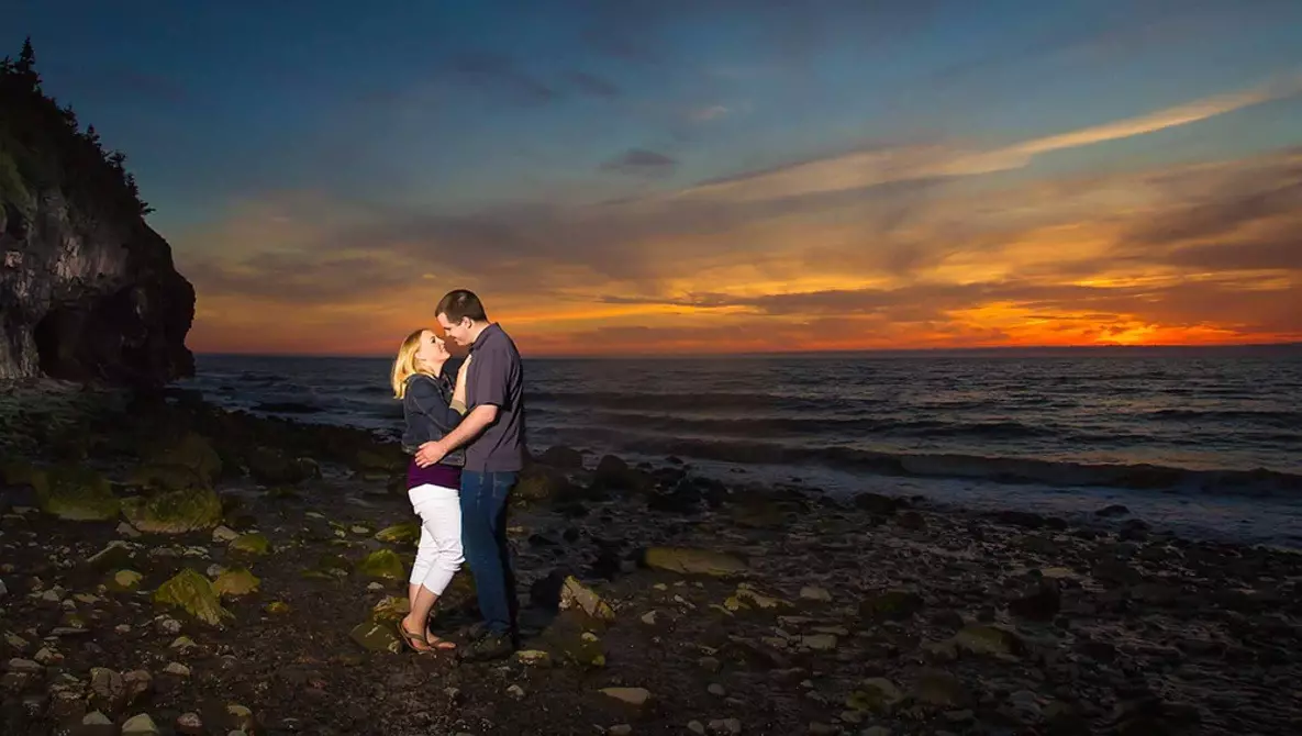 Çifti i fejuar duke u përqafuar në plazhin shkëmbor në perëndim të diellit